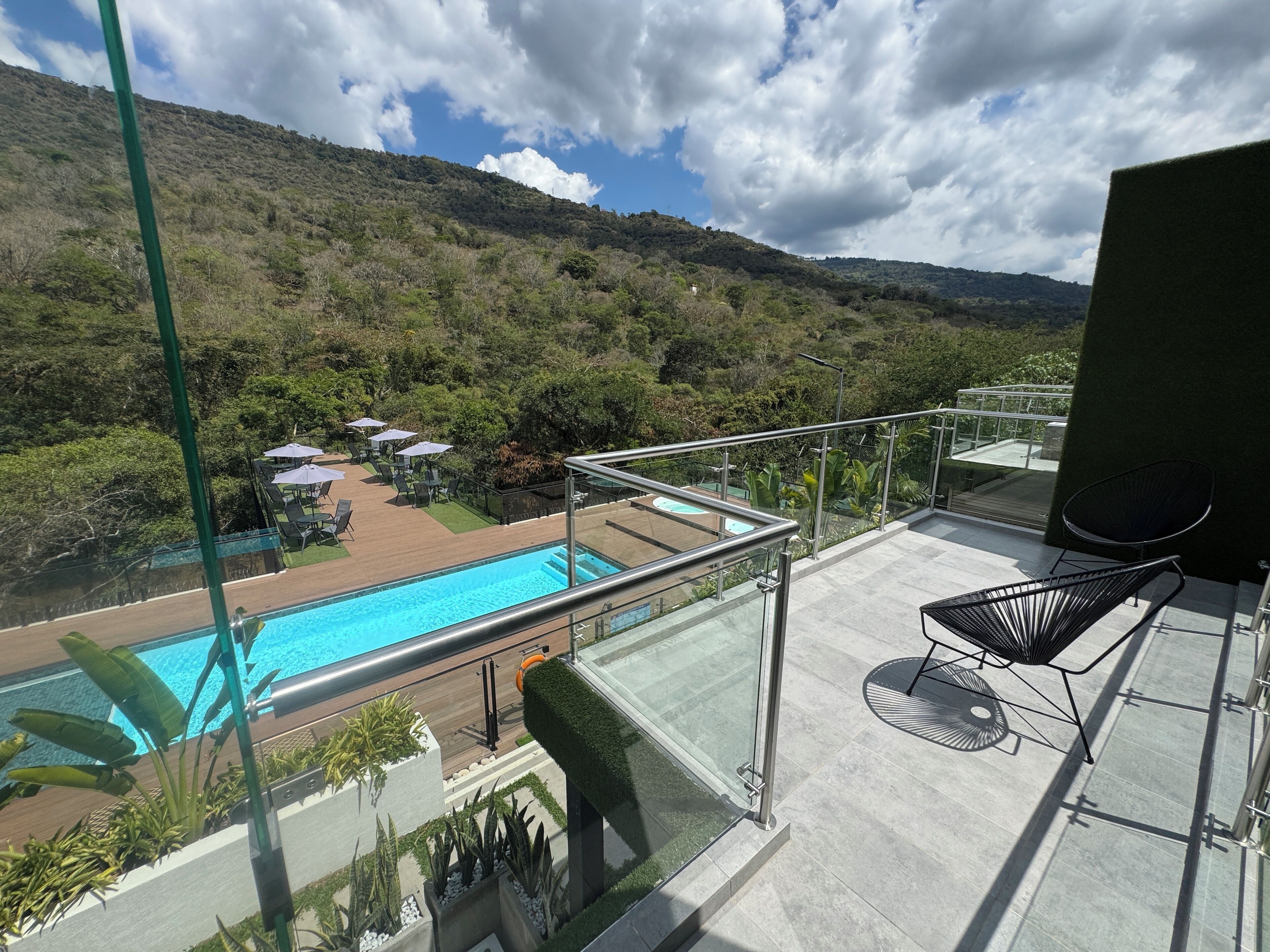 Piscina no terraço com guarda-corpo de vidro, área de estar externa e vista para um morro coberto de vegetação.