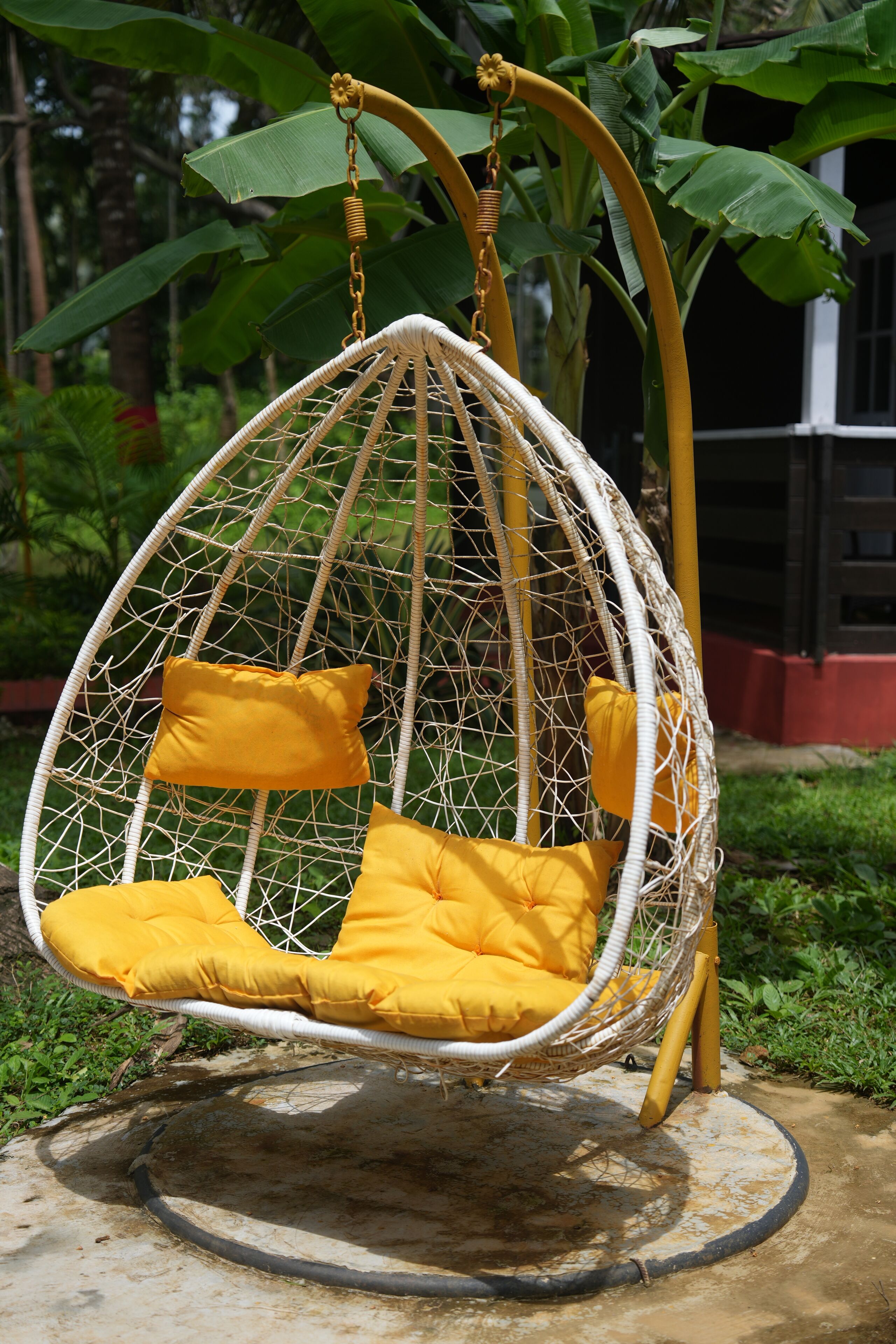 Uma cadeira de balanço branca de vime suspensa, com almofadas amarelas, em um jardim.