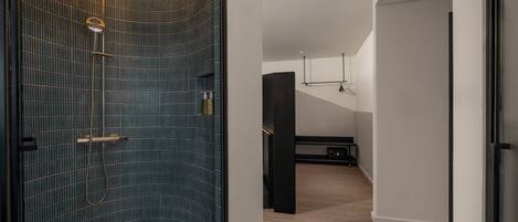 Um banheiro moderno com parede de chuveiro curva, piso escuro e corredor que leva a uma área de cozinha.
