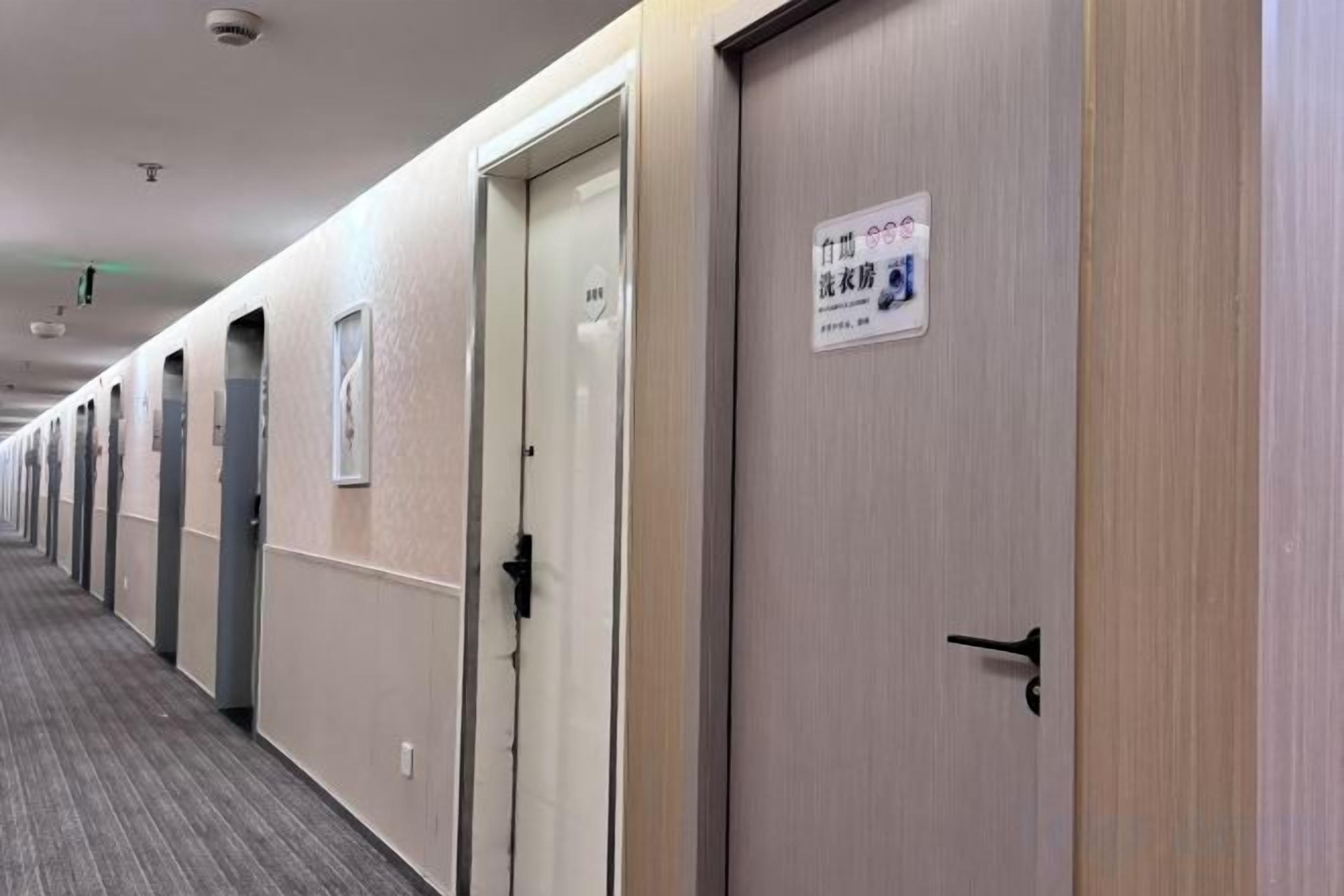 Corredor de hotel com portas, algumas com placas indicativas, e piso coberto por carpete.