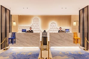 Recepção de hotel com balcão de mármore, dois recepcionistas e carpete estampado.