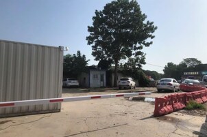 Uma área de estacionamento com entrada por portão, uma árvore e um edifício ao fundo.