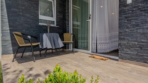 Um pátio com duas cadeiras de vime, uma mesa com toalha de tecido cinza e uma porta de vidro que dá para uma área coberta.