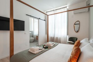 Quarto de hotel com cama, uma cadeira, um espelho e um banheiro visível por uma porta aberta.