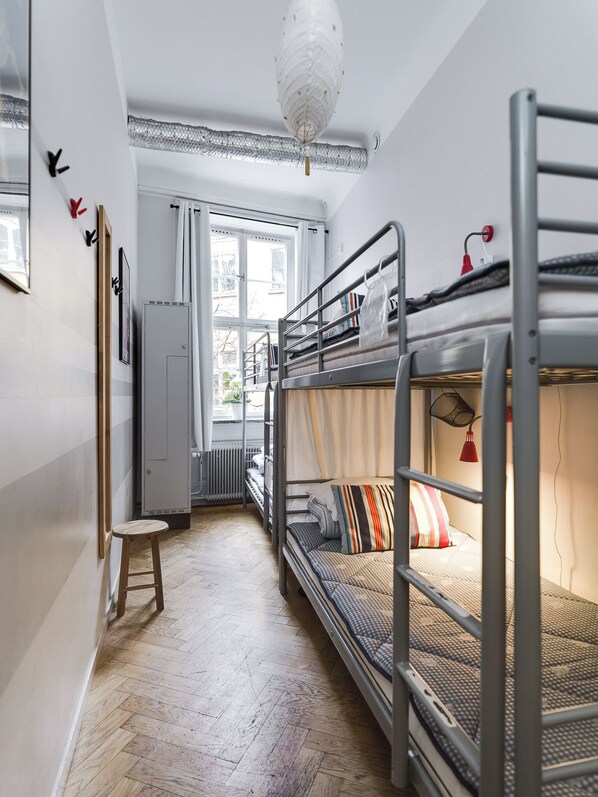 Um dormitório com beliches, um radiador, uma janela com cortinas e um banquinho de madeira.