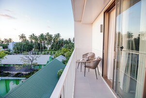 Uma varanda com duas cadeiras e uma mesa, com vista para um edifício com telhado verde e palmeiras.