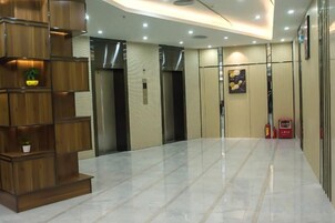 Um hall de hotel com piso de mármore, estantes de madeira com plantas e dois elevadores.