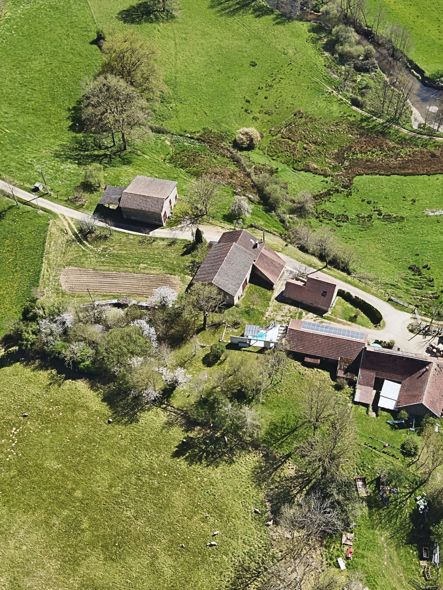 Vista aérea de uma propriedade rural com vários edifícios, incluindo casas e celeiros, rodeada por campos verdes e um pequeno riacho.