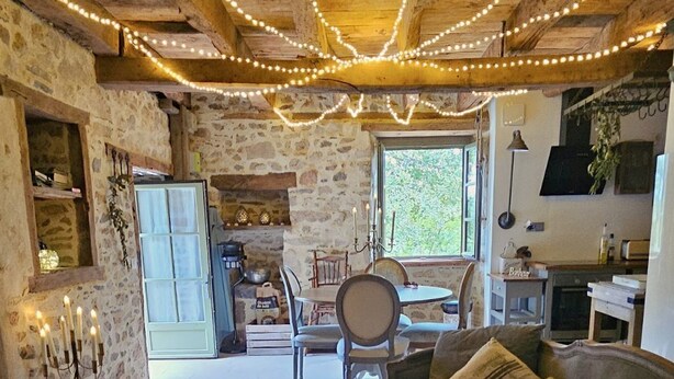 Uma sala de estar rústica com vigas de madeira expostas, paredes de pedra, uma lareira de pedra, uma área de refeições com mesa e cadeiras, e um espaço de estar aconchegante com sofá e mesa de centro com velas.