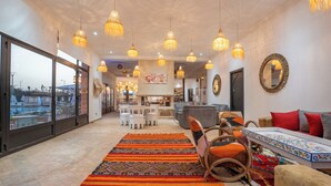 Uma sala ampla com uma grande janela, uma área de refeições com cadeiras e uma mesa, e um espaço de estar com sofás e um tapete estampado.