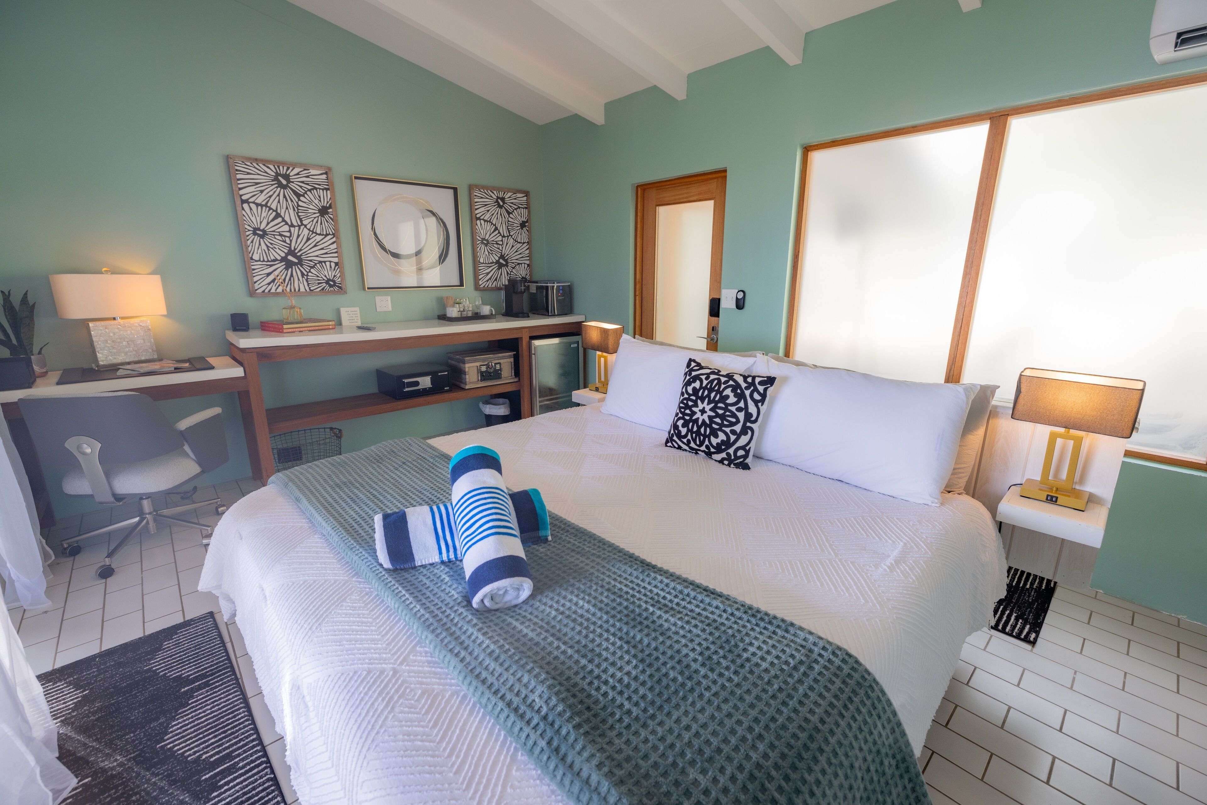 Quarto de hotel bem arrumado com uma cama grande, uma secretária com computador, uma cadeira e uma kitchenette pequena.