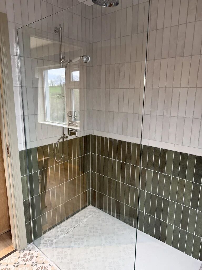 Um chuveiro com porta de vidro, uma janela acima e chão revestido com azulejos.