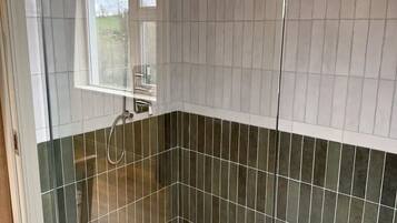 Um chuveiro com porta de vidro, uma janela acima e chão revestido com azulejos.