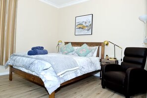 Quarto com cama, almofadas, cadeira, mesa de cabeceira, abajur e um quadro na parede.