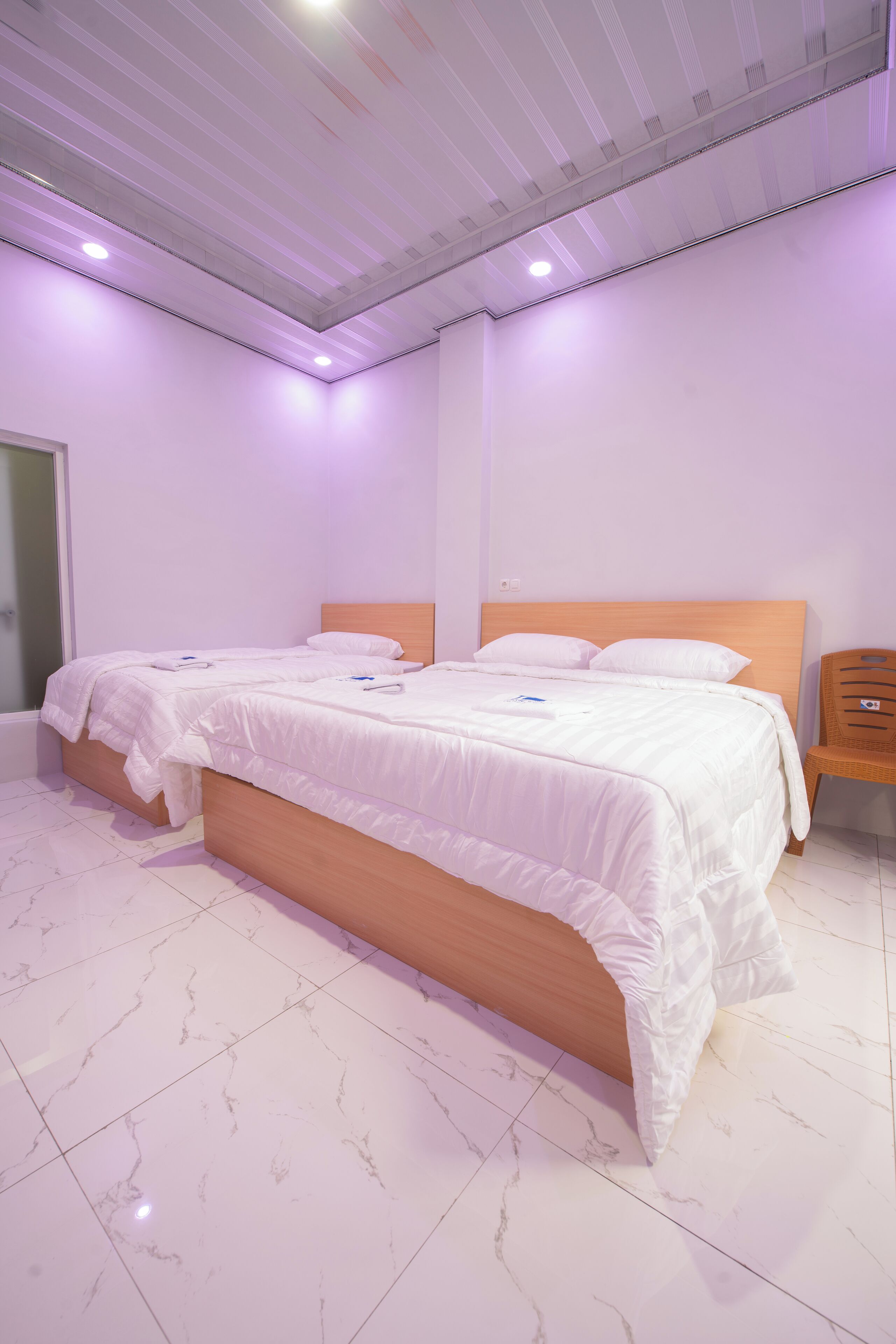 Quarto com duas camas, cabeceira de madeira, uma cadeira branca e banheiro com porta de vidro.