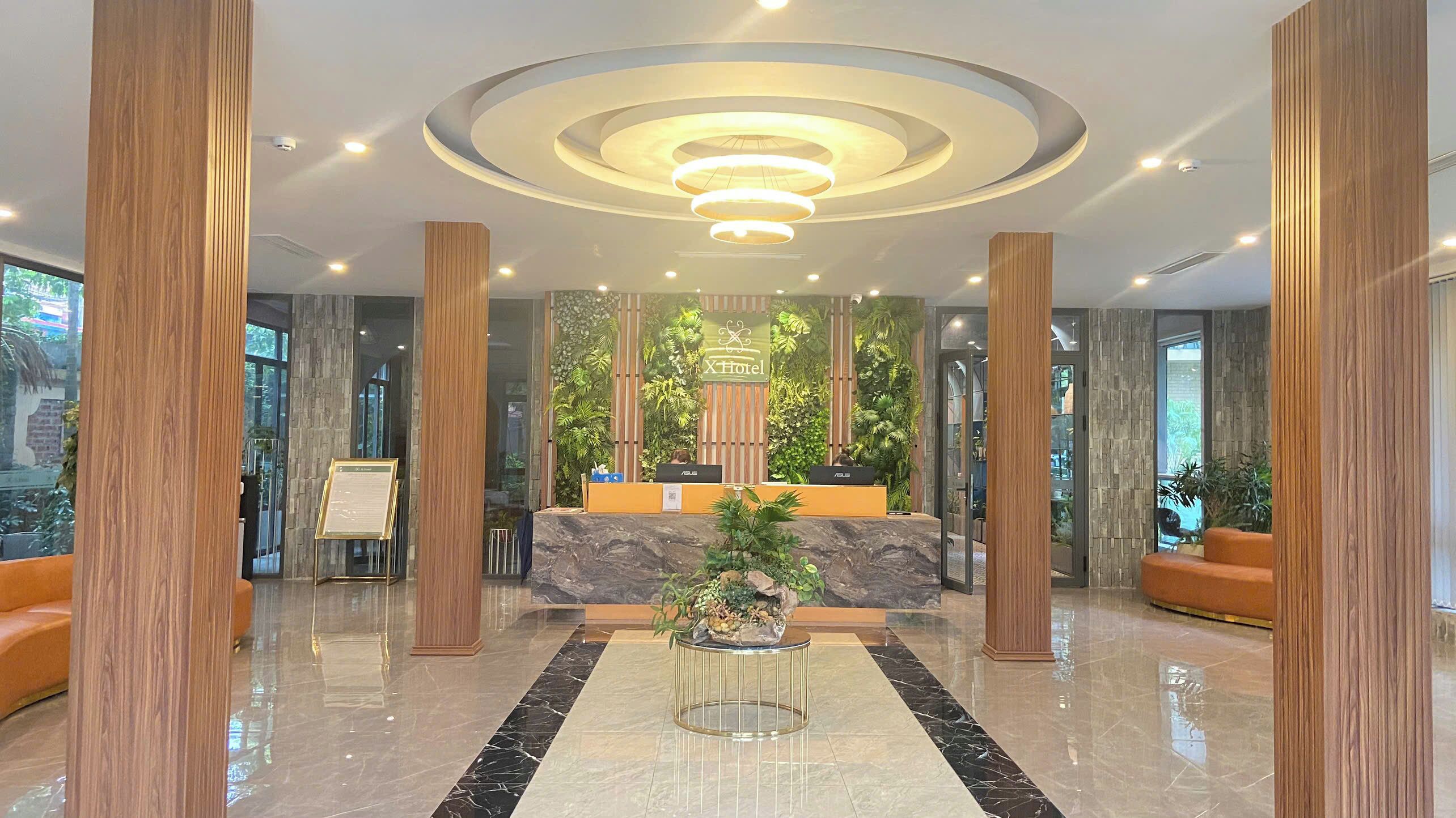 Uma área de recepção de hotel com design moderno, com piso de mármore, colunas de madeira e um balcão central de recepção com um arranjo de plantas.