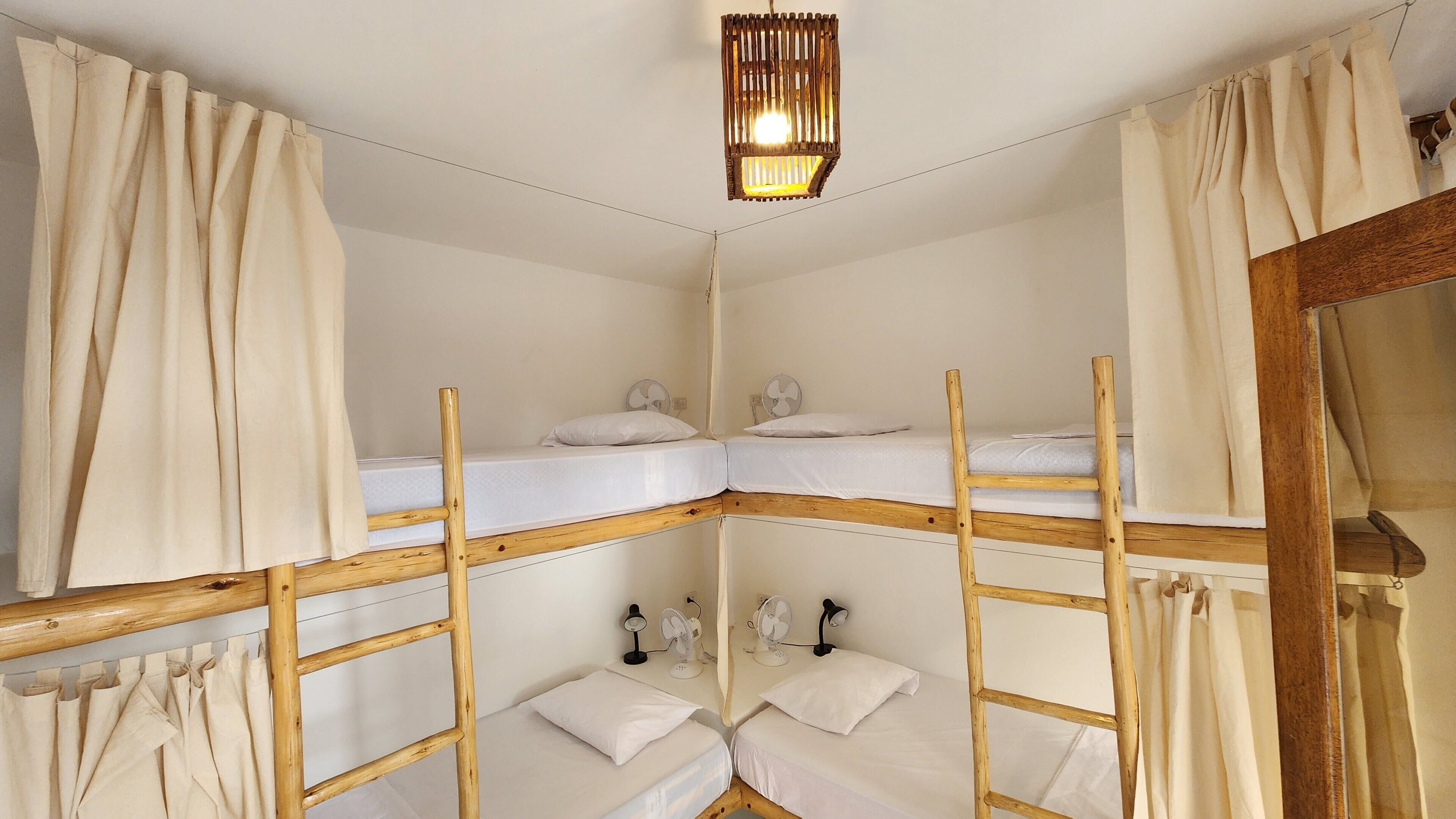 Quarto com beliches, grades de madeira, roupa de cama branca e ventilador de teto.