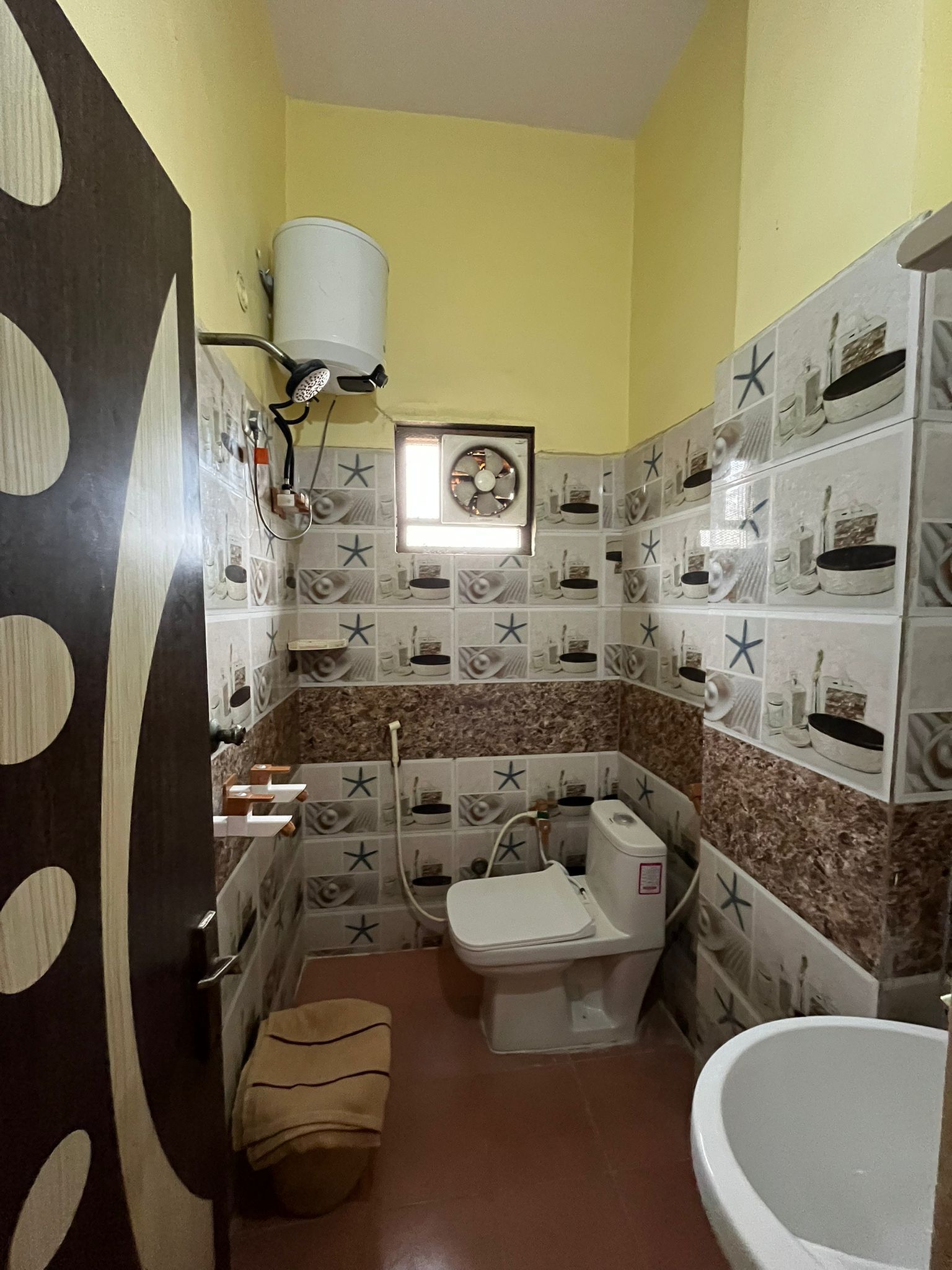 Um banheiro compacto com vaso sanitário, pia e banheira. As paredes são revestidas com um padrão de estrelas-do-mar, e há uma prateleira com diversos artigos de higiene pessoal.