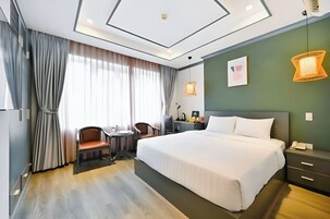 Quarto de hotel com uma cama grande, uma escrivaninha com cadeira, uma mesinha pequena, um telefone e uma luminária na parede.