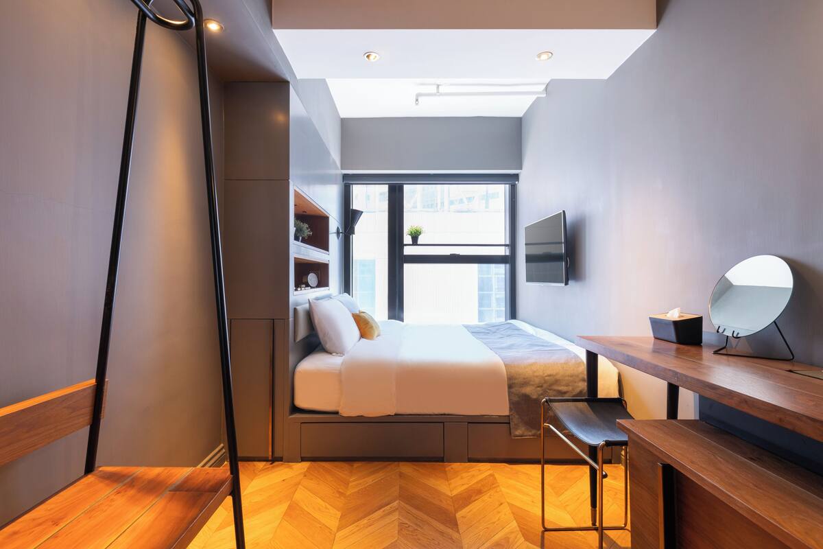 Quarto moderno com cama, escrivaninha, cadeira, televisão e janela.