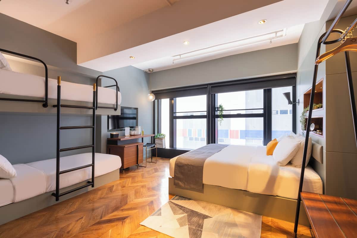 Quarto de hotel moderno com beliches, uma escrivaninha com computador e janelas amplas com vista para a cidade.