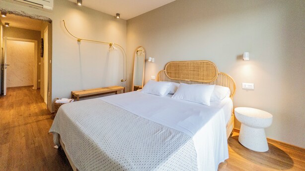 Quarto moderno com cama grande, mesa de cabeceira, um banco, vaso sanitário e espelho.