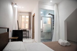 Quarto de hotel com cama, escrivaninha, cadeira, área de chuveiro e janela com cortinas.