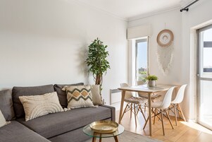 Uma sala de estar moderna com um sofá cinzento, uma mesa de centro de vidro e um espaço de refeições com uma mesa e cadeiras brancas.