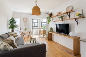 Uma sala de estar moderna com sofá, televisão, prateleiras de madeira com plantas e um espaço de jantar com cadeiras e mesa.