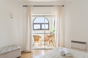 Quarto com cama, um banco, radiador e vista para uma varanda com cadeiras e uma mesa.