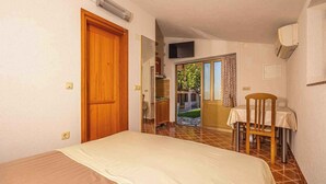 Quarto com cama, porta de madeira, mesa pequena com cadeira e vista para um jardim através de uma porta aberta.