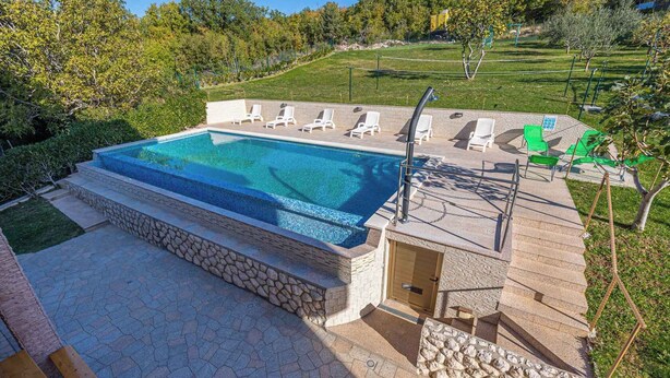 Piscina com um deck de pedra, rodeada por espreguiçadeiras e uma área gramada.