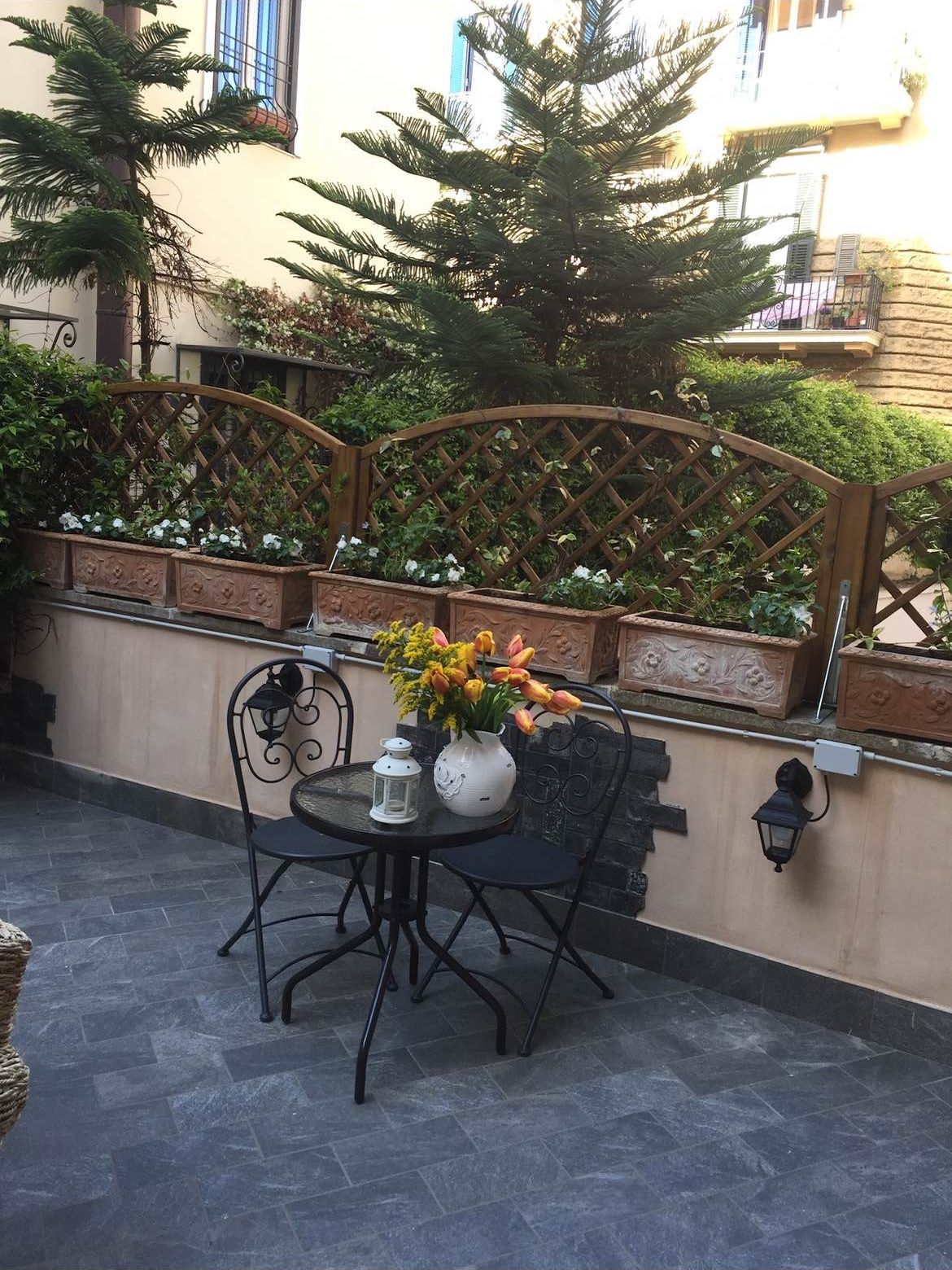 Um pátio externo com uma mesa redonda, duas cadeiras, um vaso com flores e um candeeiro.