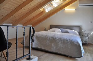 Quarto com teto inclinado, cama com cabeceira cinza, mesa de cabeceira com abajur e tapete pequeno no chão.