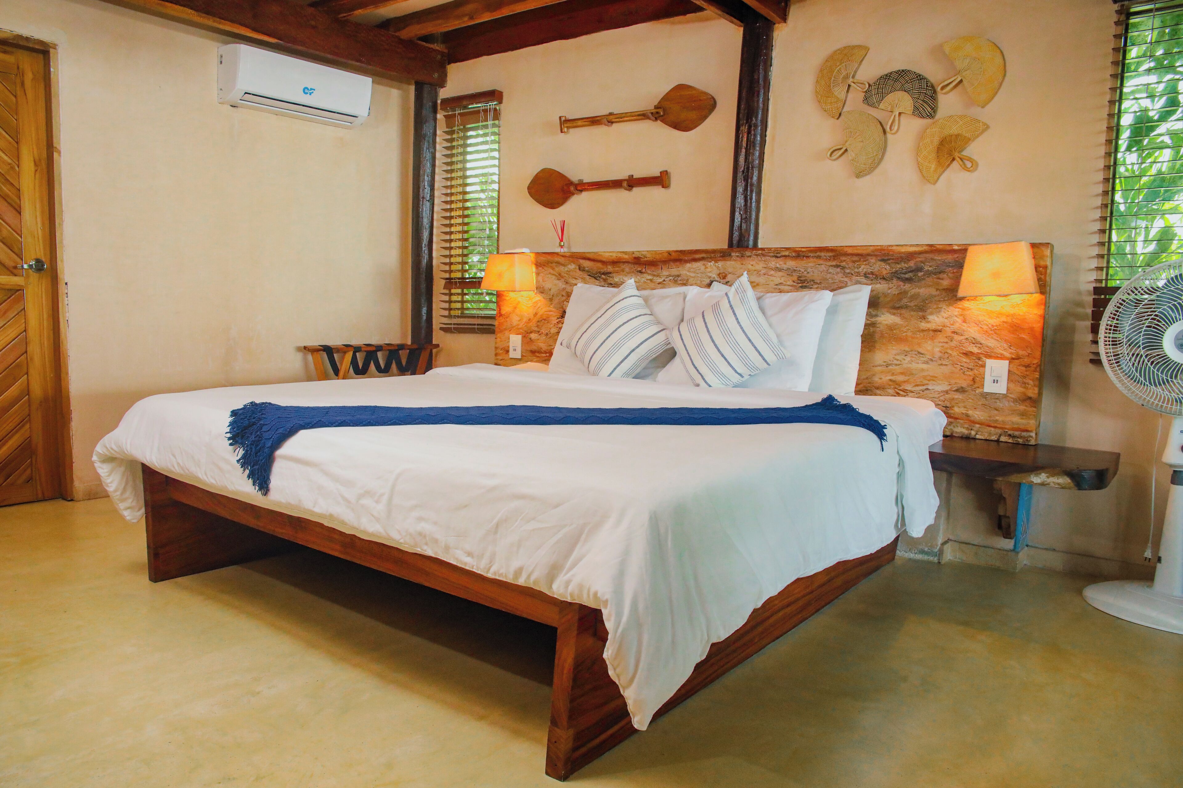 Uma cama de madeira com roupa de cama branca e um cobertor azul, um ar condicionado fixado na parede e um ventilador.