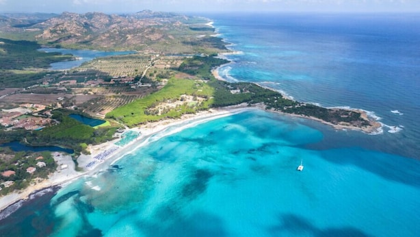 Vista aérea de uma área costeira com praia, águas azul-claras e vegetação ao redor.