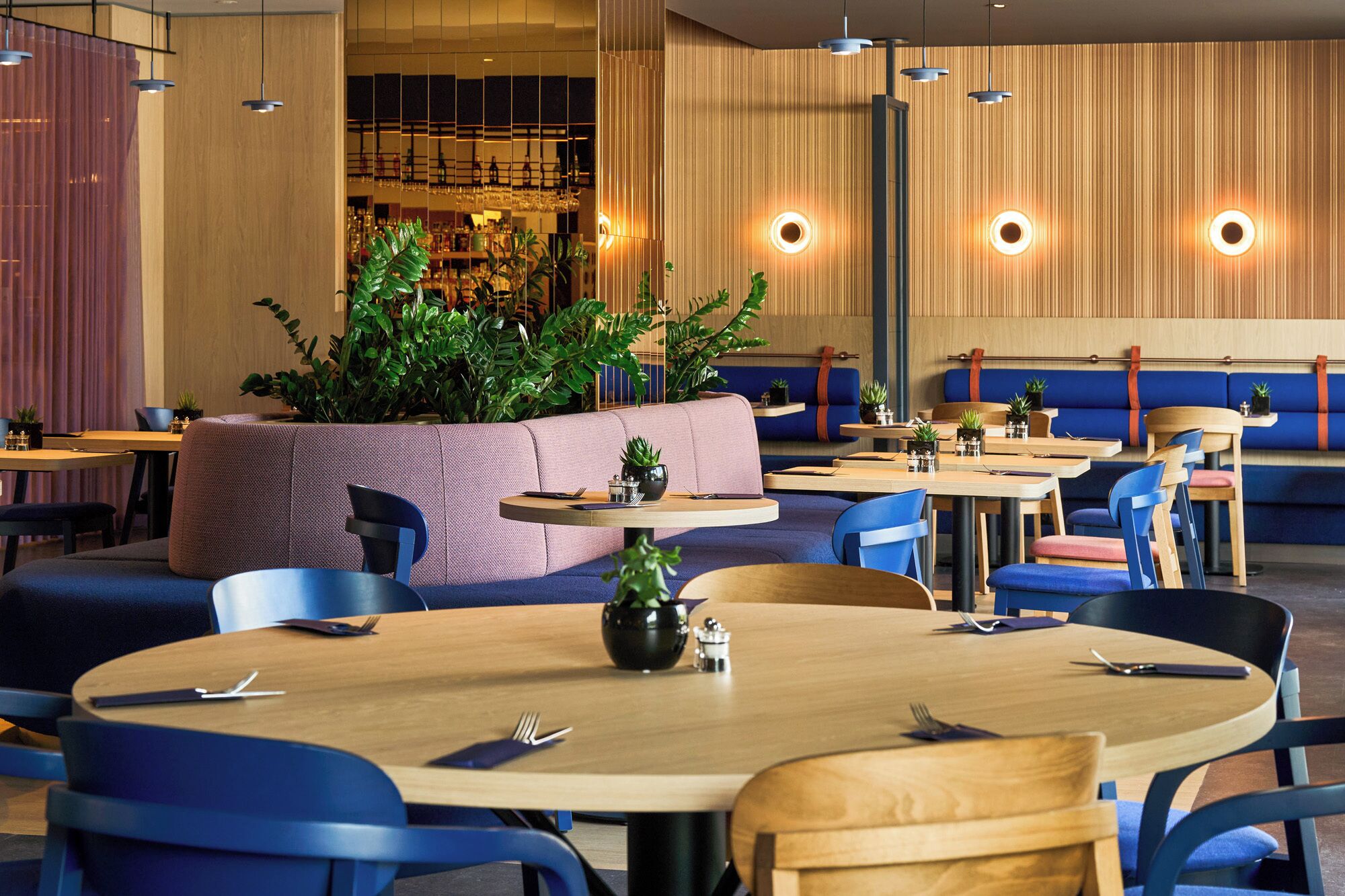 Interior de restaurante moderno com mesas redondas, cadeiras azuis e área central de estar com um banco acolchoado e plantas.