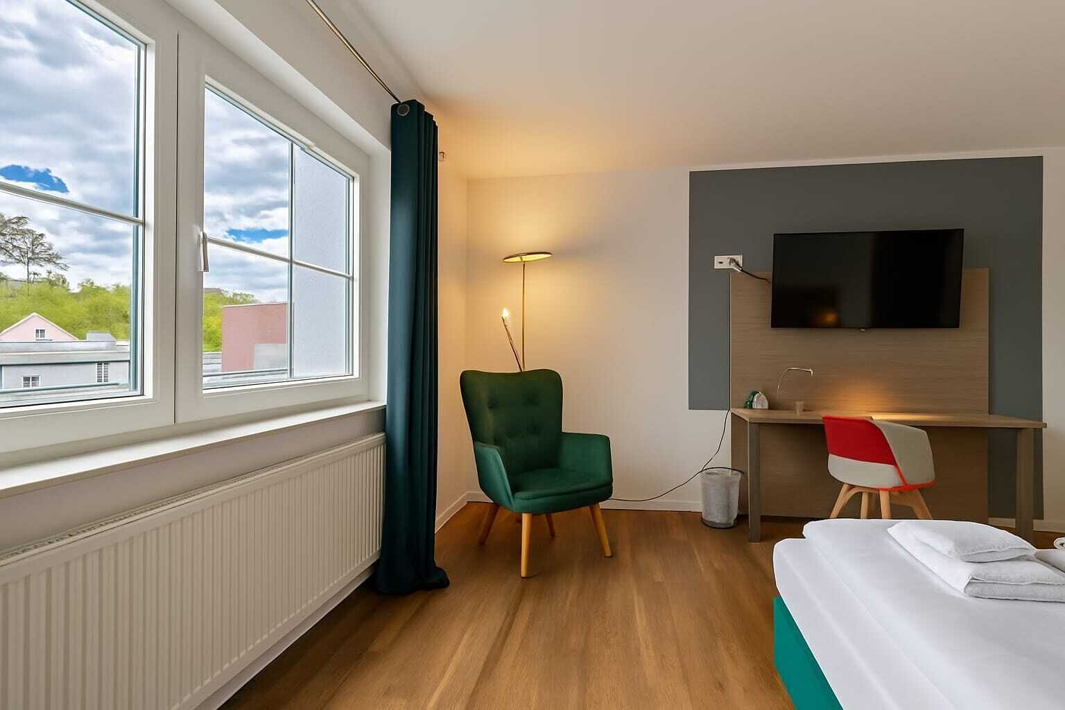 Quarto de hotel com uma cama, uma escrivaninha com televisão, uma cadeira verde e vista para edifícios lá fora.