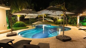 Piscina com cadeiras de sol e guarda-sóis ao redor, cercada por vegetação e palmeiras.