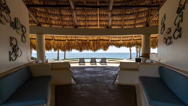 Quarto com telhado de palha, duas áreas de estar com assentos azuis e vista para o oceano.
