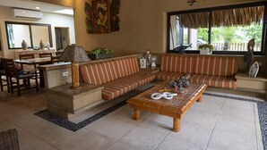 Ampla sala de estar com um sofá listrado, uma mesa de centro com objetos decorativos, uma área de jantar com cadeiras e uma grande janela com vista para áreas verdes.