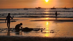 Cena de praia com pessoas e barcos ao pôr do sol.