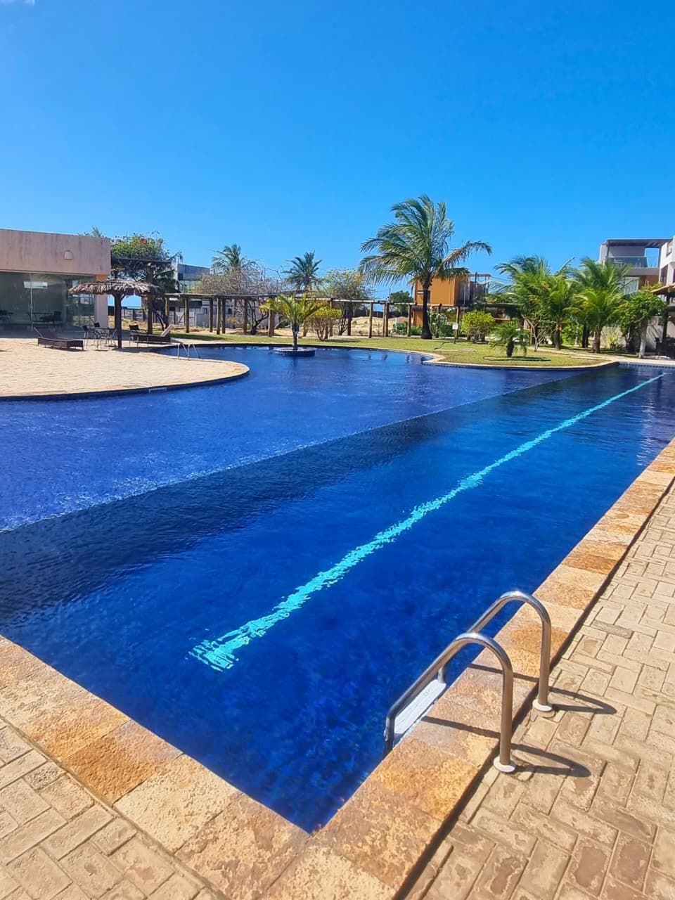 Piscina com águas azul-claras, cercada por palmeiras e um espaço coberto com assentos.
