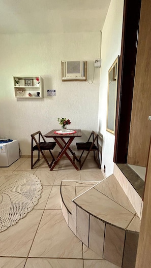 Quarto pequeno e bem iluminado, com uma mesa de jantar de madeira, duas cadeiras, uma pequena prateleira com objetos decorativos, um aparelho de ar condicionado e piso ladrilhado.