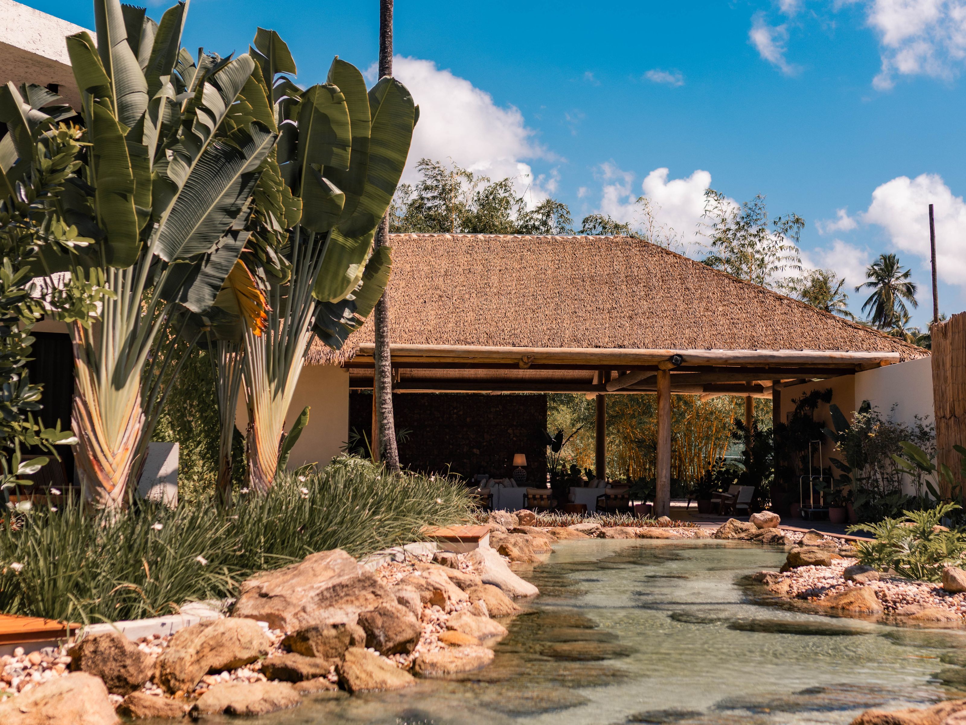 Um resort tropical com um pavilhão de palha, um espelho d'água límpido e vegetação exuberante.