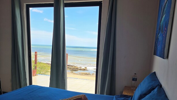 Cama com roupa de cama azul, uma toalha enrolada e um travesseiro, com vista para a praia e o oceano através de uma janela.