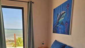 Quarto com cama, quadro, mesa de cabeceira e vista para o oceano.