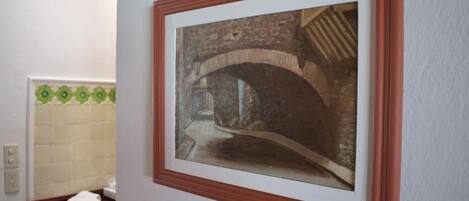 Pintura emoldurada de uma ponte de pedra arqueada em um banheiro com pia branca e toalha no balcão.