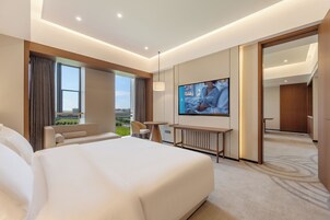 Quarto de hotel com uma cama grande, televisão, área de estar com mesa e vista para o exterior.