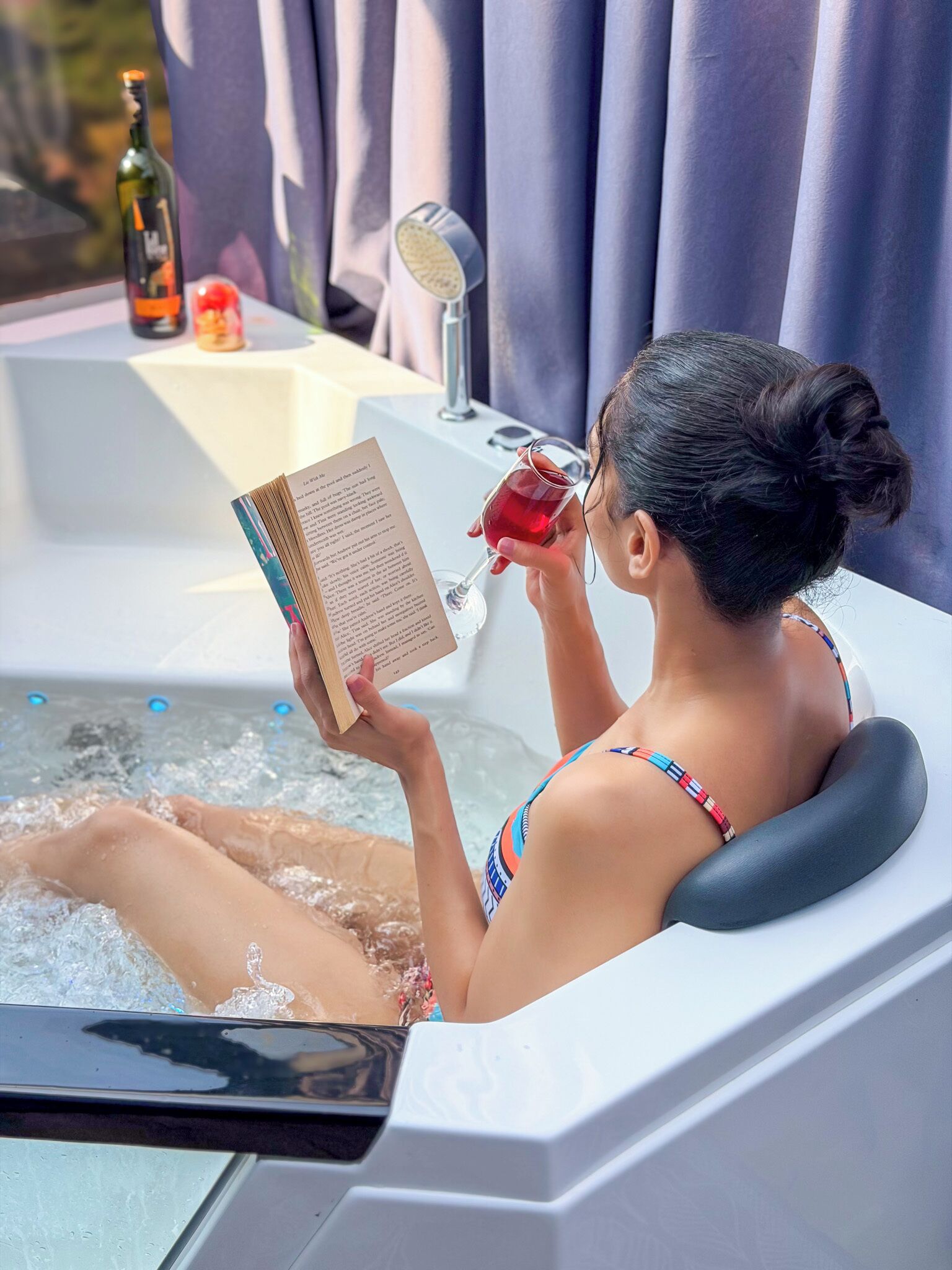 Uma pessoa na banheira lendo um livro, com uma garrafa e um copo ao lado.